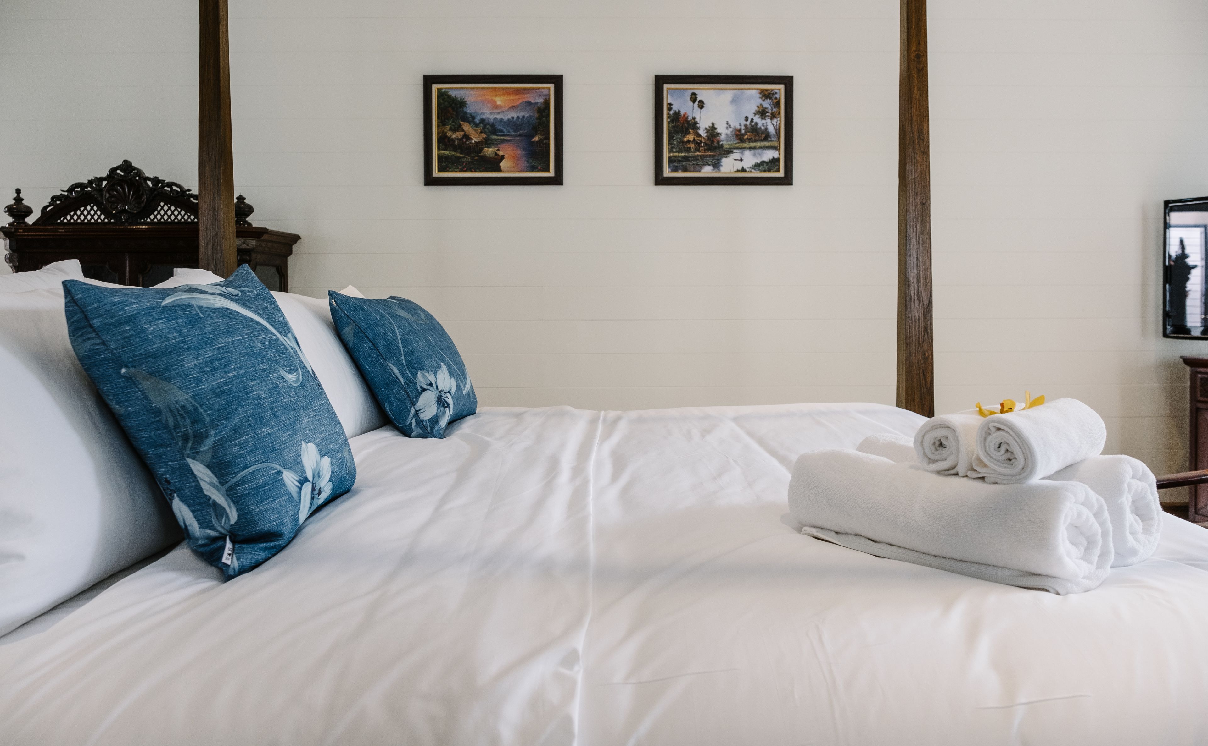 Cama bem arrumada com lençóis brancos e almofadas florais azuis, duas pinturas de paisagens emolduradas na parede e cabeceira de madeira.