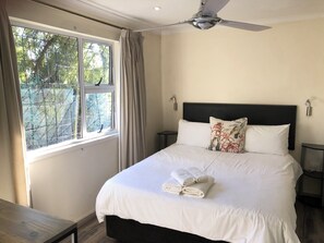 Uma cama bem arrumada com lençóis brancos e duas toalhas dobradas, um ventilador de teto, uma janela com cortinas e uma mesa de cabeceira com abajur.