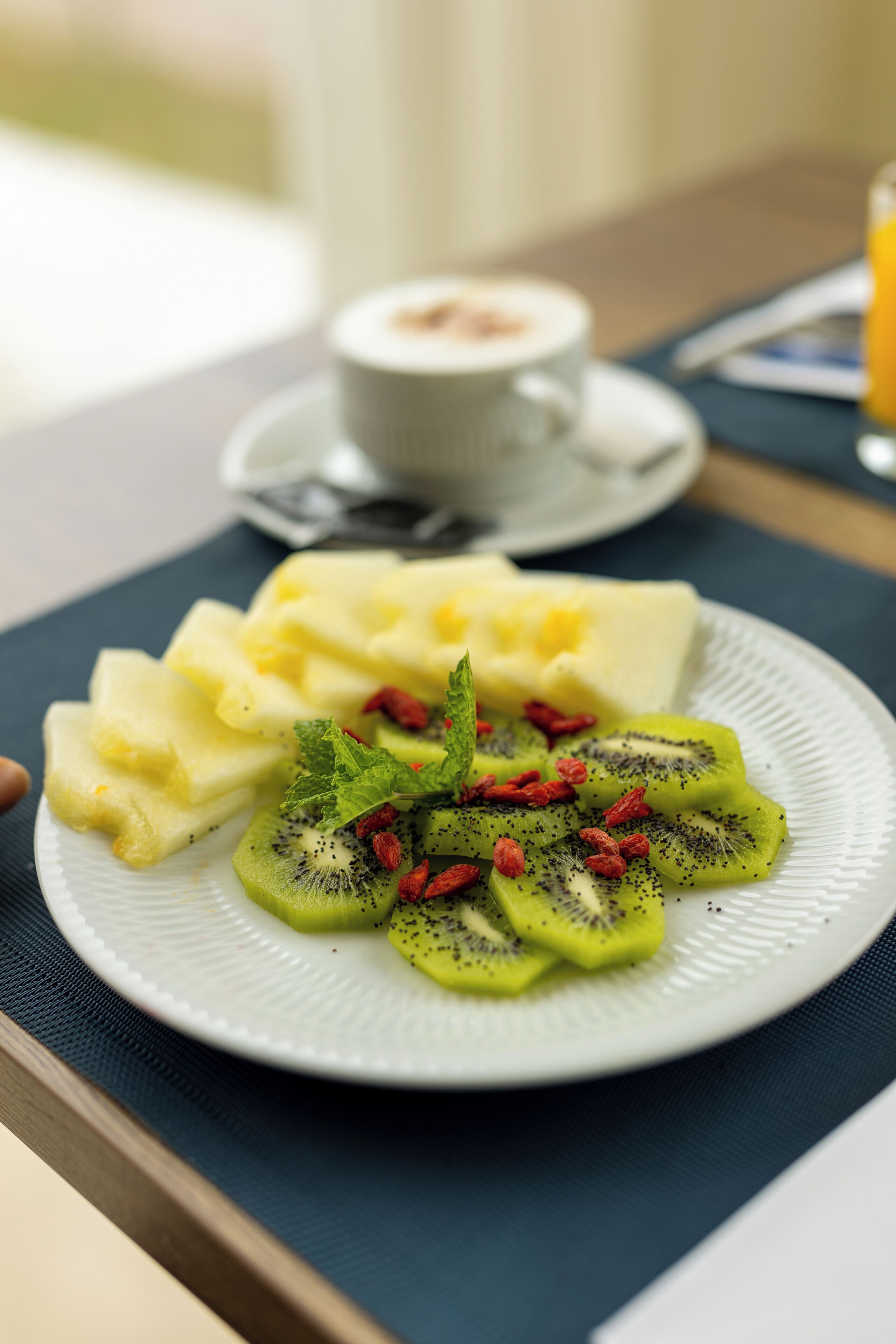 Um prato com kiwi e manga fatiados, juntamente com frutas vermelhas, uma xícara de café e um copo de suco de laranja sobre uma mesa.