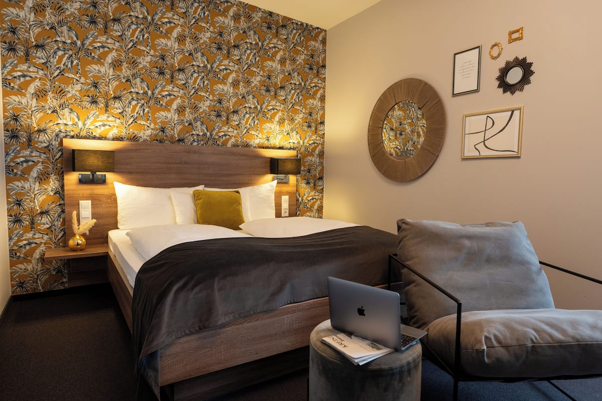 Quarto de hotel bem arrumado, com uma cama, mesa lateral com abajur, um laptop e uma cadeira.