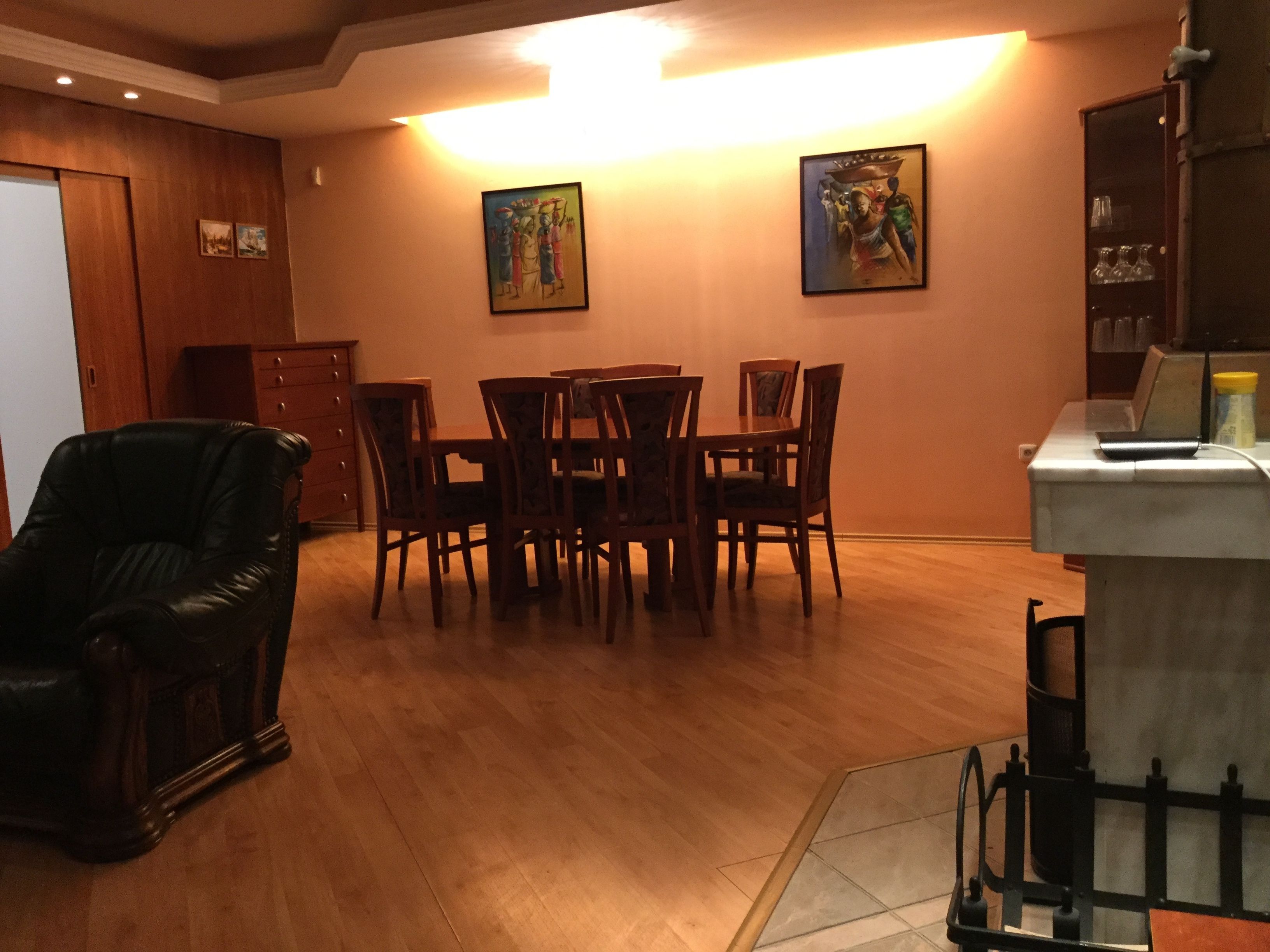 Uma área de refeições com mesa e cadeiras de madeira, uma poltrona de couro preto, armários de madeira e duas pinturas na parede.