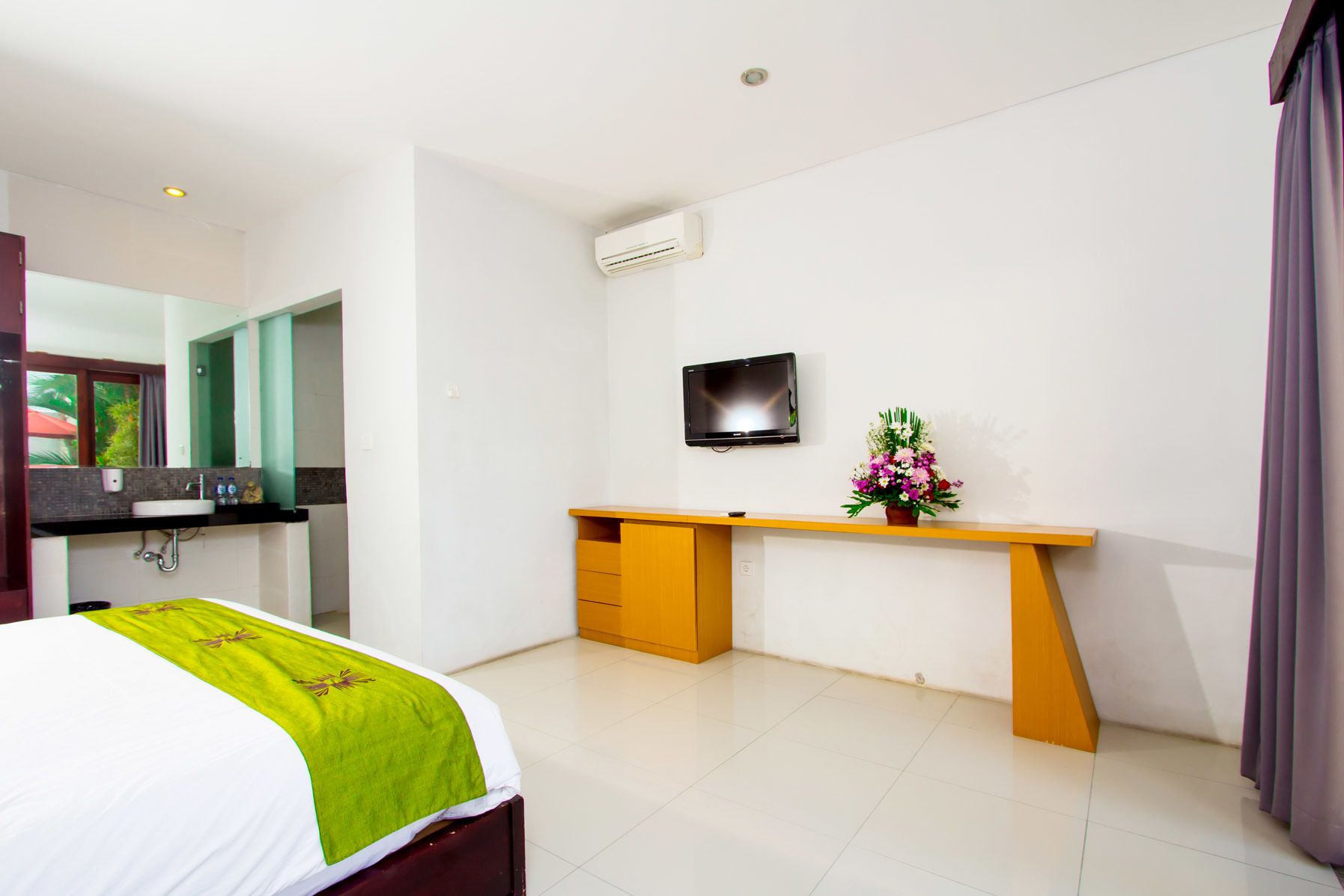 Quarto de hotel com cama, escrivaninha, televisão e banheiro visível por uma porta aberta.