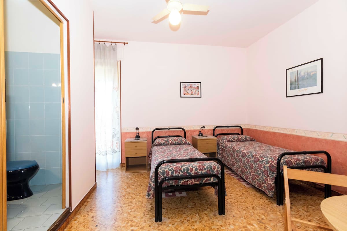 Quarto com duas camas, uma mesa de madeira, ventoinha no teto, quadro emoldurado na parede e banheiro com vaso sanitário e pia.