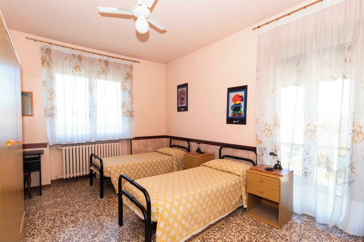 Quarto com duas camas, uma escrivaninha, uma cadeira, radiador e ventoinha no teto.