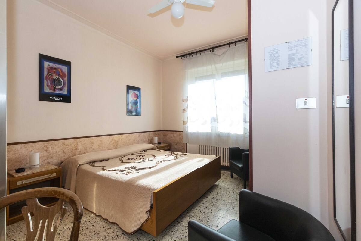Quarto com cama, cadeira, mesa de cabeceira, escrivaninha e ventoinha no teto.