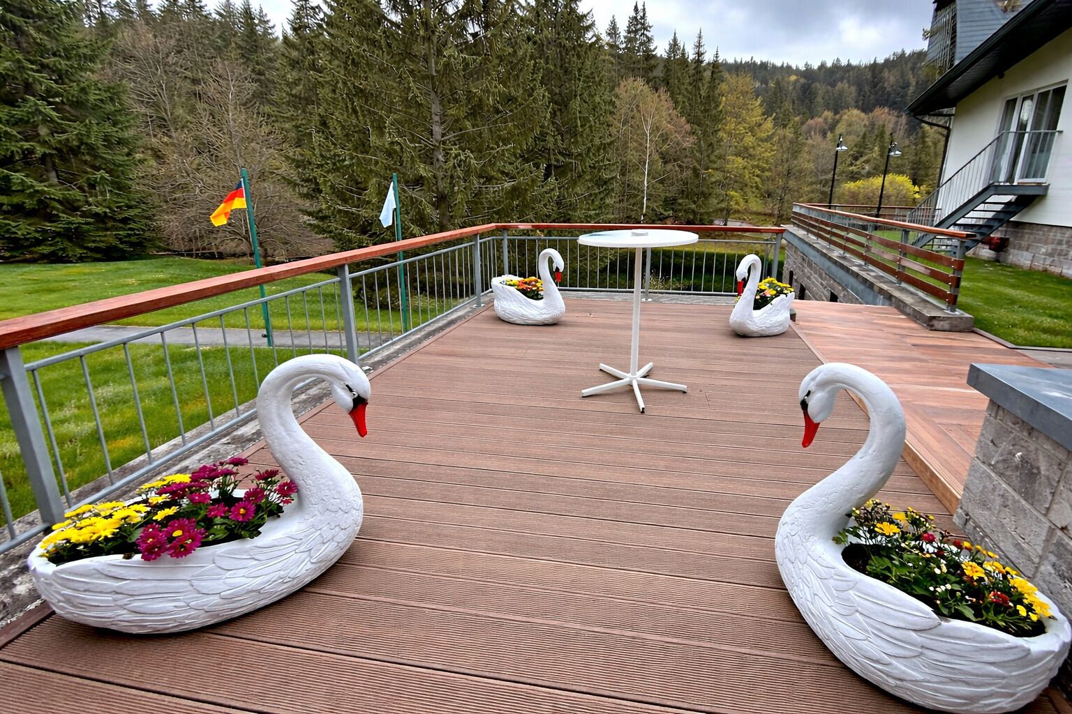Um deck de madeira com dois vasos em formato de cisne contendo flores, uma mesa redonda e um corrimão.