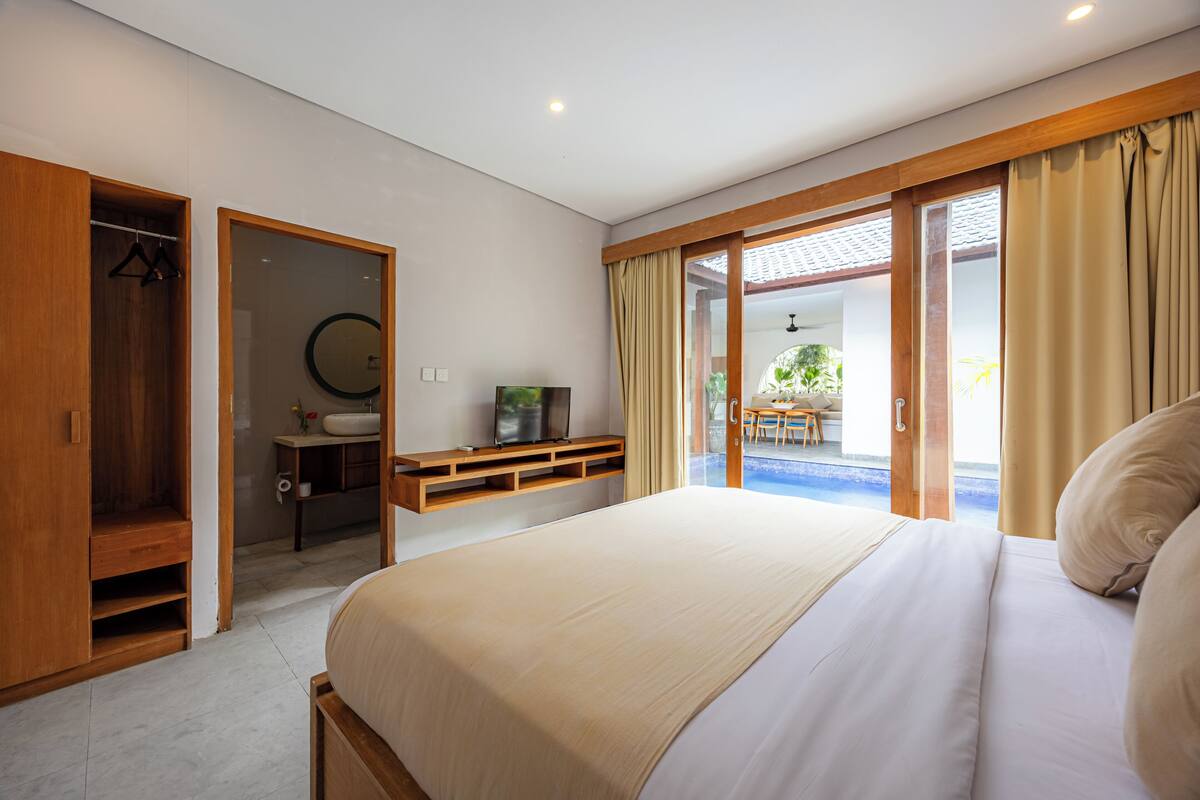 Quarto com cama, armário de madeira, televisão e banheiro visível por uma porta aberta.
