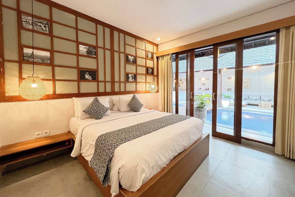 Quarto com uma cama grande, cabeceira de madeira, parede com quadros e vista para uma piscina através de portas de vidro.