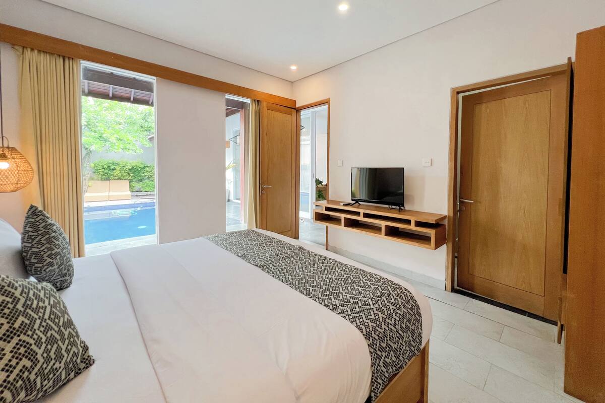 Quarto com cama, TV em prateleira de madeira, porta de vidro deslizante e vista para uma piscina.