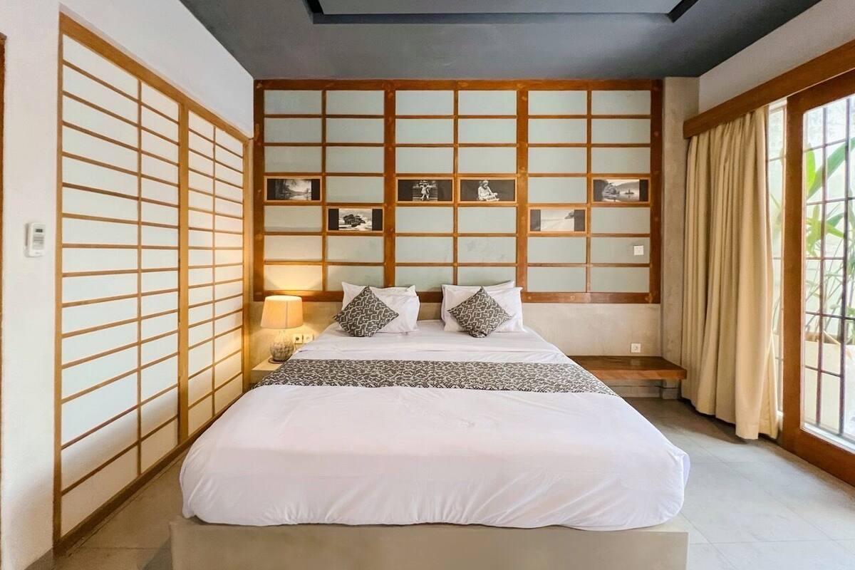 Quarto de hotel com uma cama grande, cabeceira de madeira com arte, mesa de cabeceira com abajur e vista pela janela com cortinas.