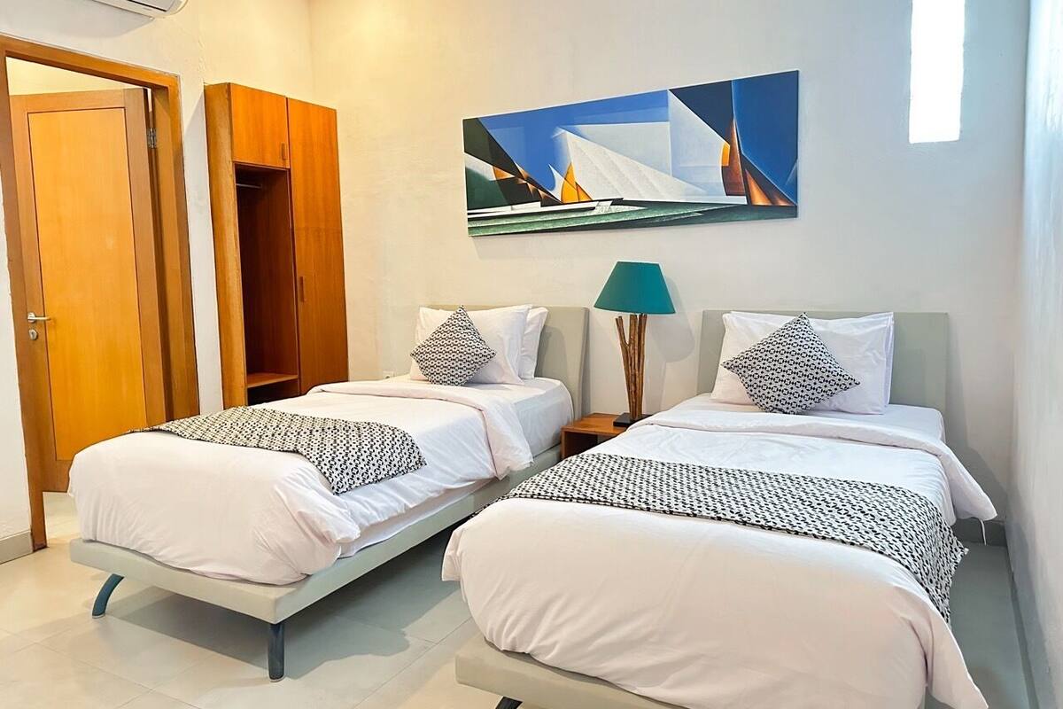 Quarto de hotel com duas camas, guarda-roupa de madeira, pintura de um veleiro e abajur teal.