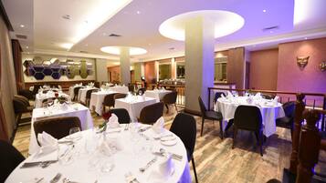 Uma sala de jantar de hotel com mesas redondas preparadas para refeições, com toalhas de mesa brancas, talheres bem dispostos e copos.
