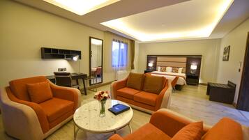 Quarto de hotel com dois sofás laranja, uma mesa de centro redonda, uma cama com cabeceira, uma escrivaninha e um espelho na parede.