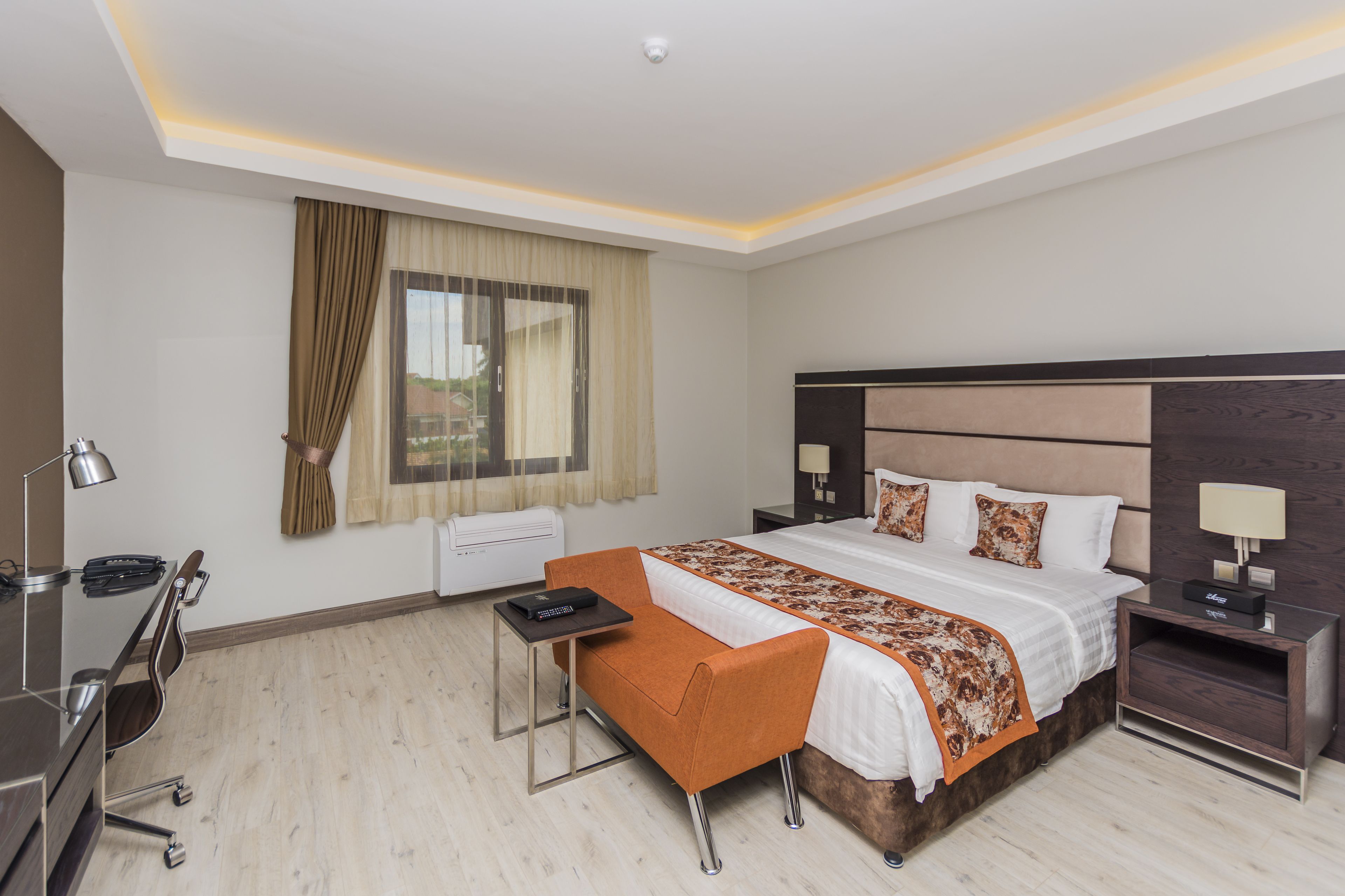 Quarto de hotel com uma cama grande, uma escrivaninha, uma cadeira, uma mesinha de cabeceira e uma janela com cortinas.