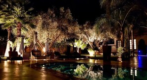 Uma área de piscina ao ar livre à noite, com espreguiçadeiras e palmeiras, iluminada por iluminação ambiente.