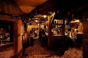 Uma área de bar acolhedora, com iluminação suave, piso de pedra e vigas de madeira, com balcão e assentos para os hóspedes.