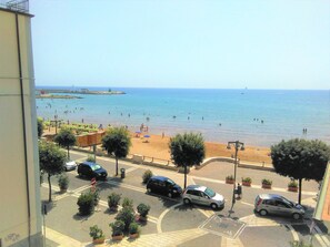 Vista da praia a partir de uma varanda, com céu claro, areal arenoso e vários carros estacionados.