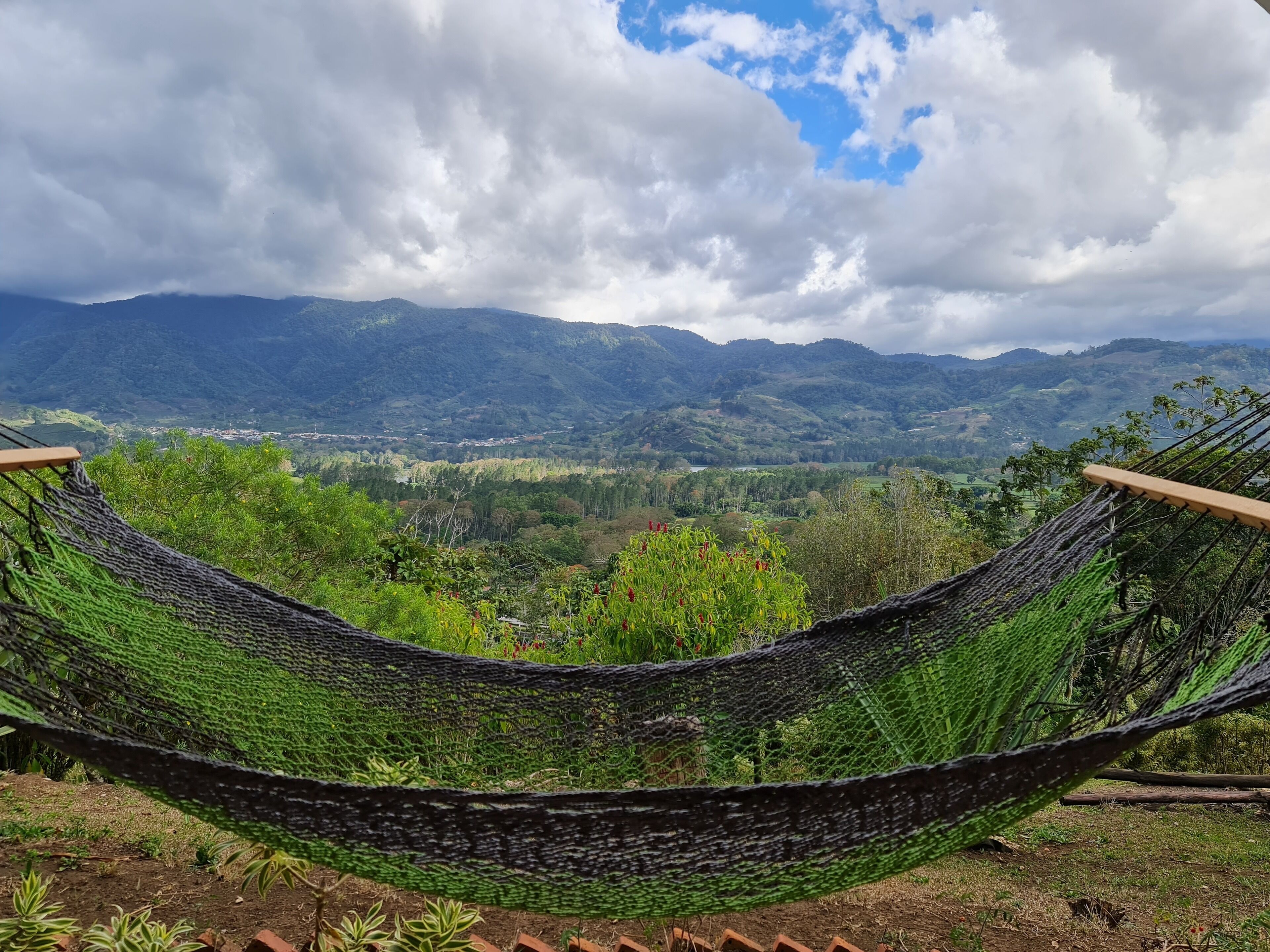 Uma rede montada em um terraço com vista para uma paisagem montanhosa, com vegetação exuberante e colinas ao longe.