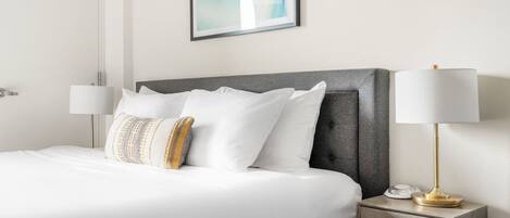 Cama bem arrumada com lençóis brancos e cabeceira cinza, ladeada por duas mesas de cabeceira com abajures e relógio.