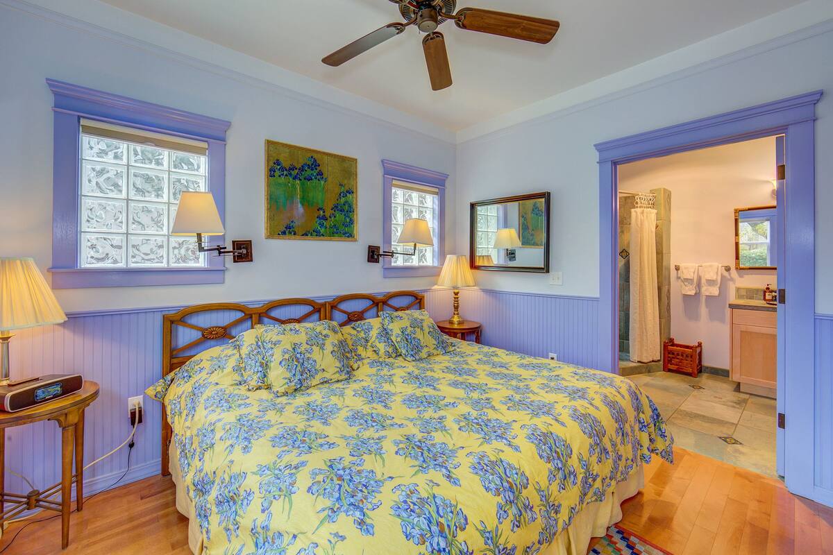 Quarto com uma cama grande, duas mesas de cabeceira com abajures, um ventilador de teto e um banheiro visível por uma porta aberta.