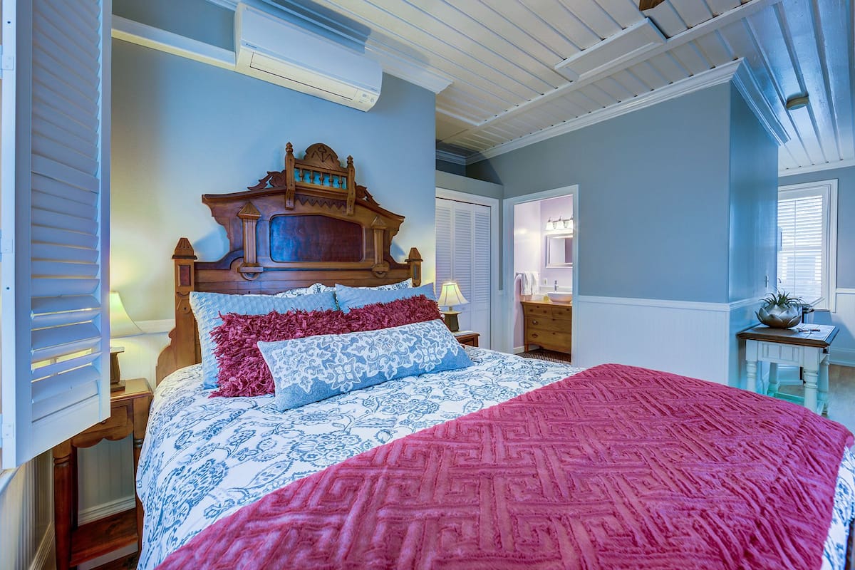 Quarto com uma cama grande, cabeceira de madeira, mesa de cabeceira, janela com persianas e banheiro visível através de uma porta aberta.