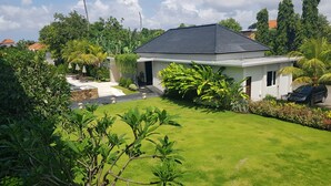 Uma casa moderna com um gramado bem cuidado, uma área de pátio e diversas plantas tropicais.