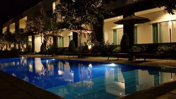 Uma área de piscina à noite, com espreguiçadeiras e guarda-sóis, cercada por um edifício com amplas janelas e fachada bem iluminada.