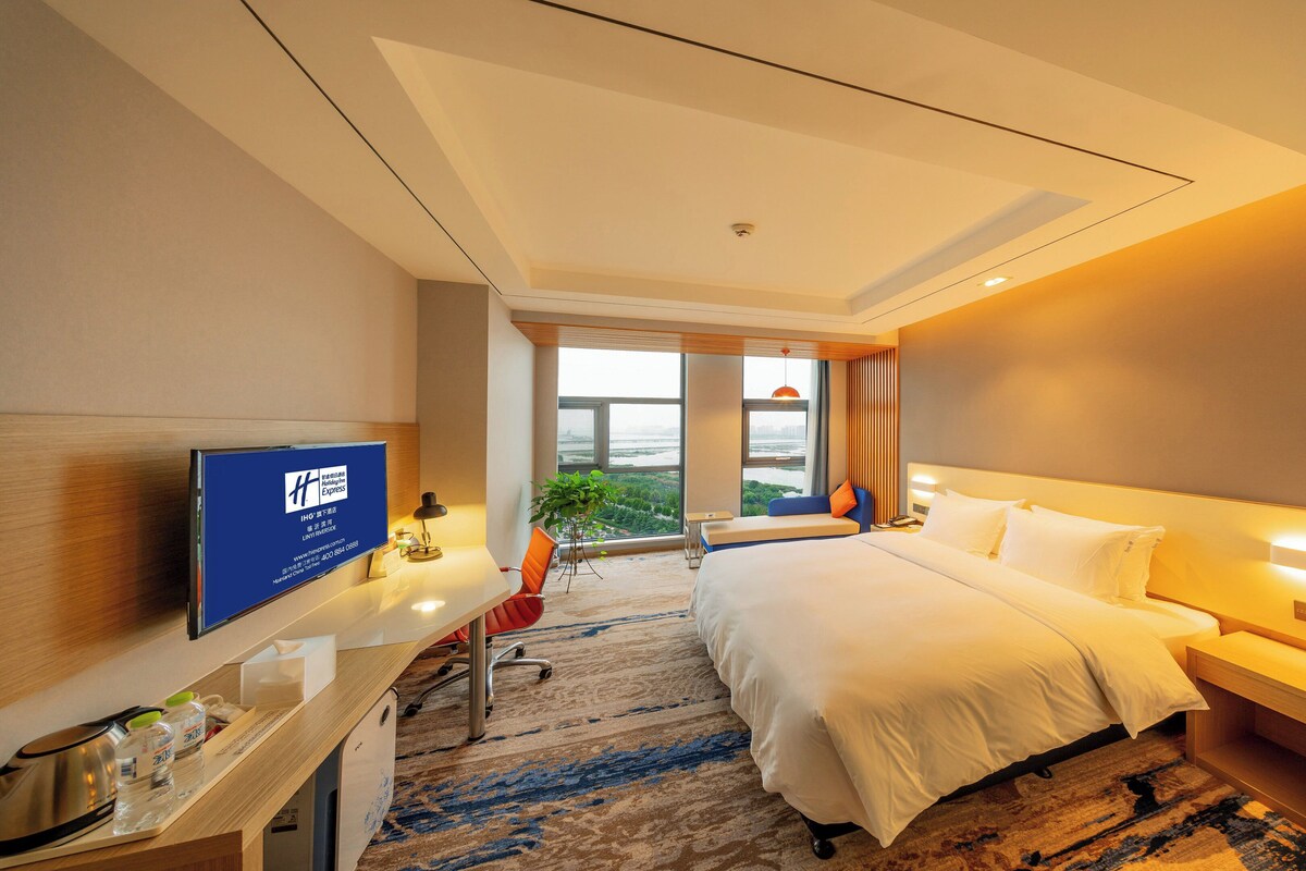 Quarto de hotel com uma cama, uma escrivaninha com um computador, uma televisão e uma janela grande com vista.