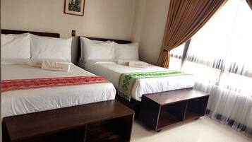 Quarto de hotel com duas camas, cada uma com roupa de cama e almofadas com a etiqueta "VERANDA". Há um banco de madeira em frente às camas, e uma janela com cortinas leves.