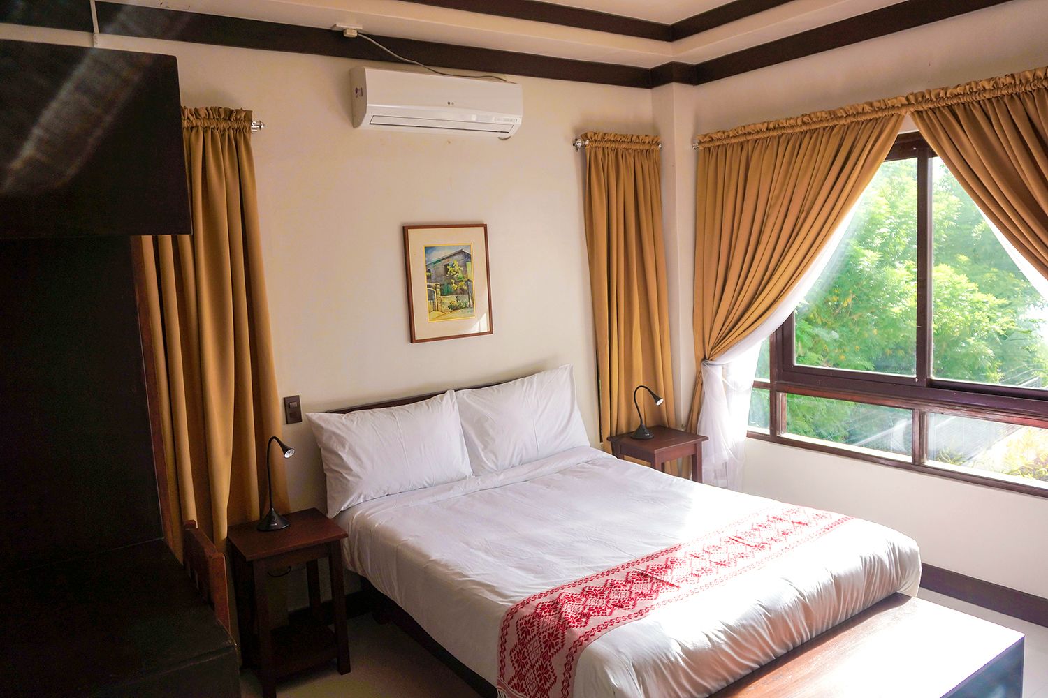 Quarto de hotel com cama, mesa de cabeceira, luminária, quadro pendurado na parede e janela com cortinas.