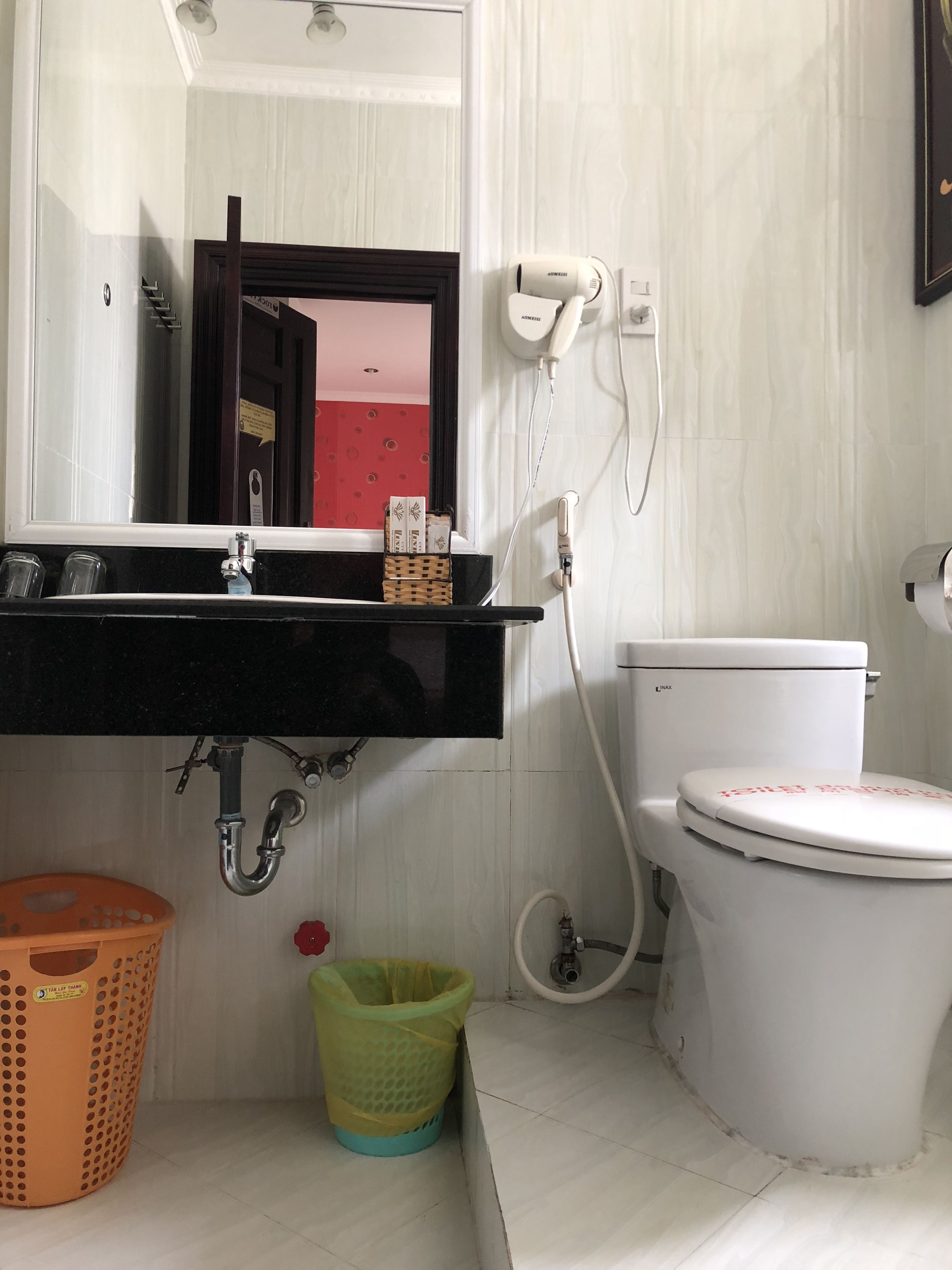 Casa de banho com sanita branca, lavatório preto com espelho em cima, secador de cabelo na parede e um contentor de lixo.