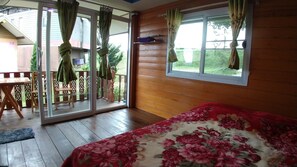 Quarto com piso de madeira, cama com colcha florida, porta de vidro deslizante para área externa com mesa e cadeiras, e janela com cortinas.