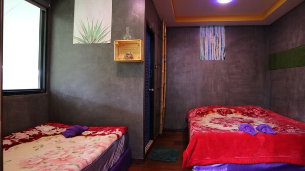 Quarto com duas camas, uma pequena prateleira de madeira com um vaso, uma planta suspensa na parede e uma porta com moldura de bambu.