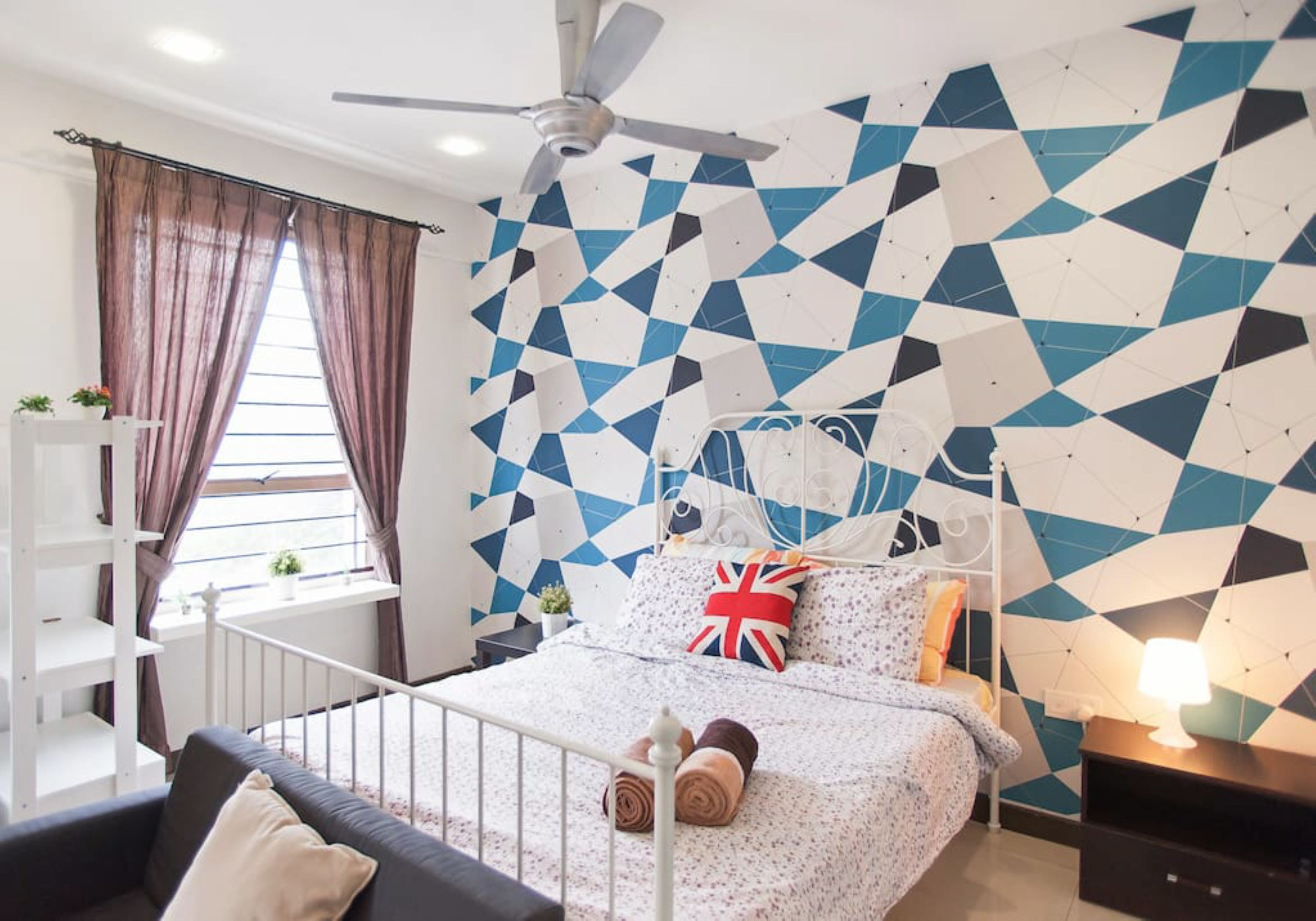 Quarto com parede estampada em padrão geométrico, cama com cabeceira branca, ventilador de teto e janela com cortinas.