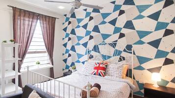 Quarto com parede estampada em padrão geométrico, cama com cabeceira branca, ventilador de teto e janela com cortinas.
