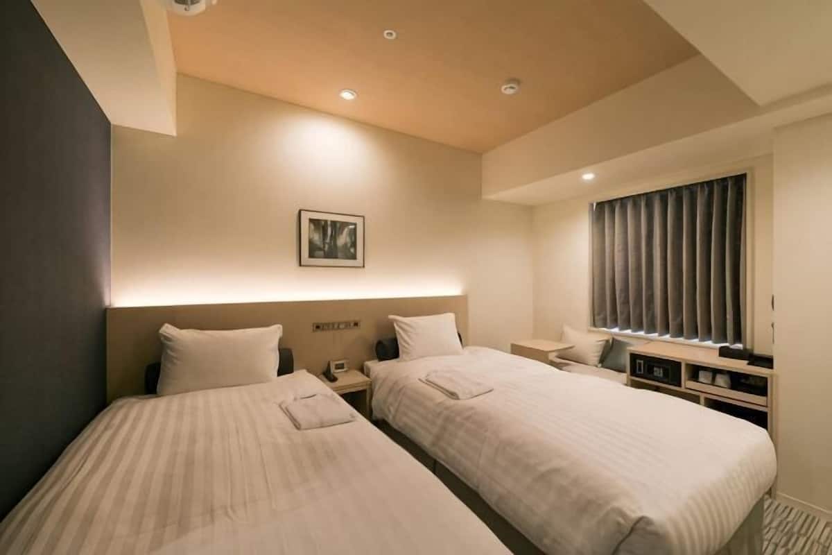 Quarto de hotel com duas camas, uma mesa de cabeceira, cortinas numa janela e um quadro pendurado na parede.