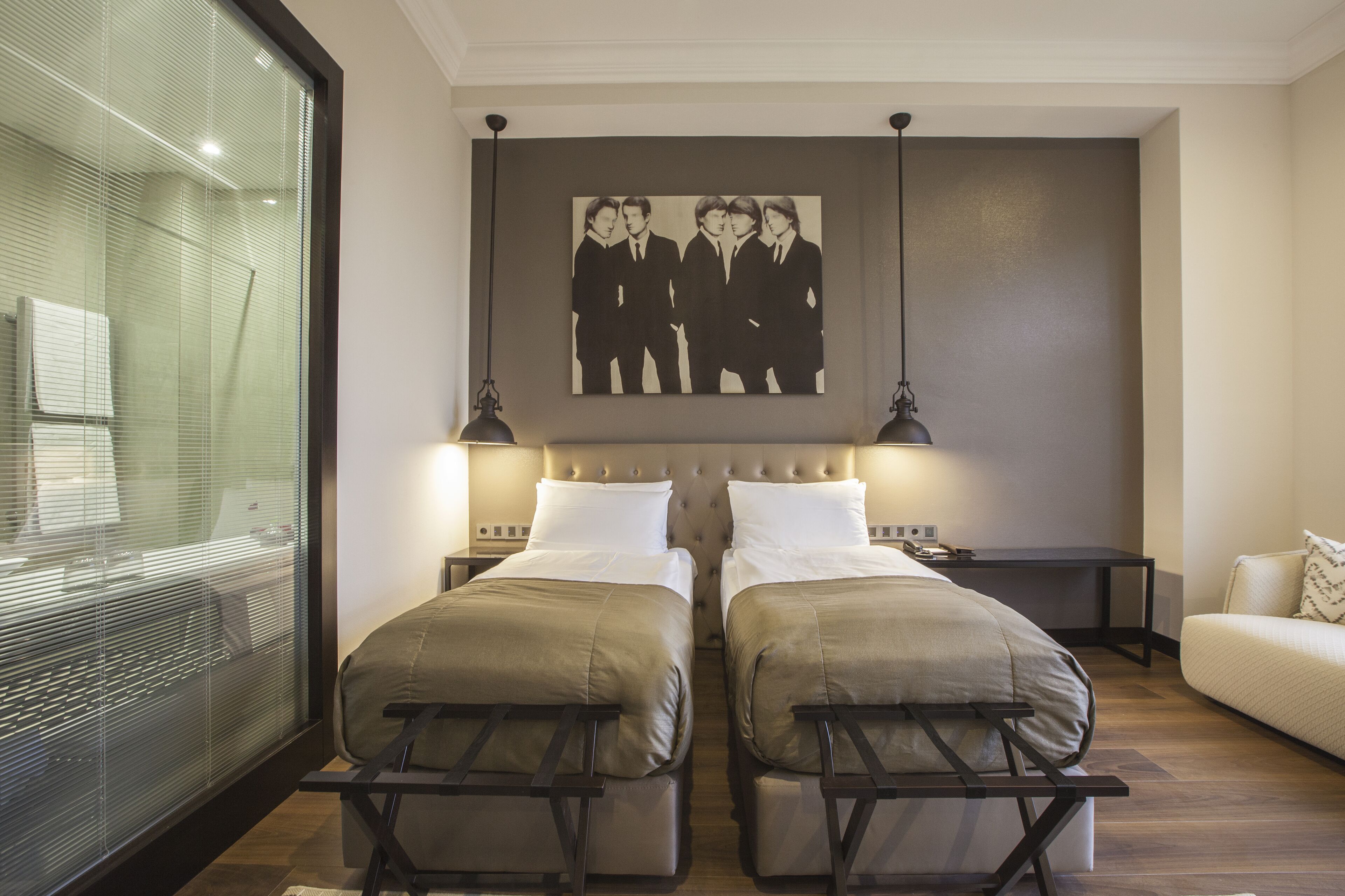 Quarto de hotel com duas camas, um sofá, um quadro na parede e uma janela com persianas.