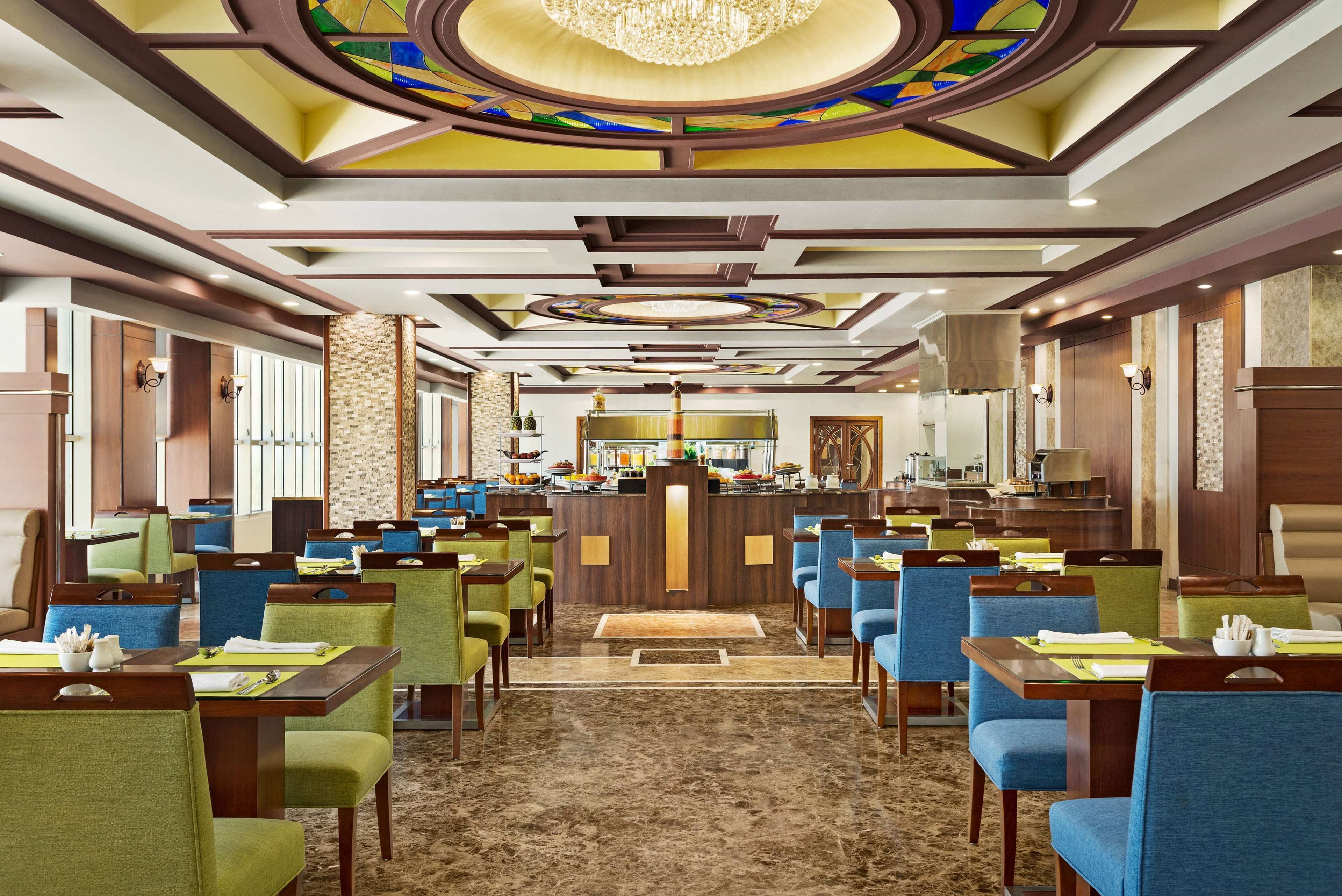 Uma sala de jantar de hotel com várias mesas e cadeiras, um balcão central e um teto estampado.