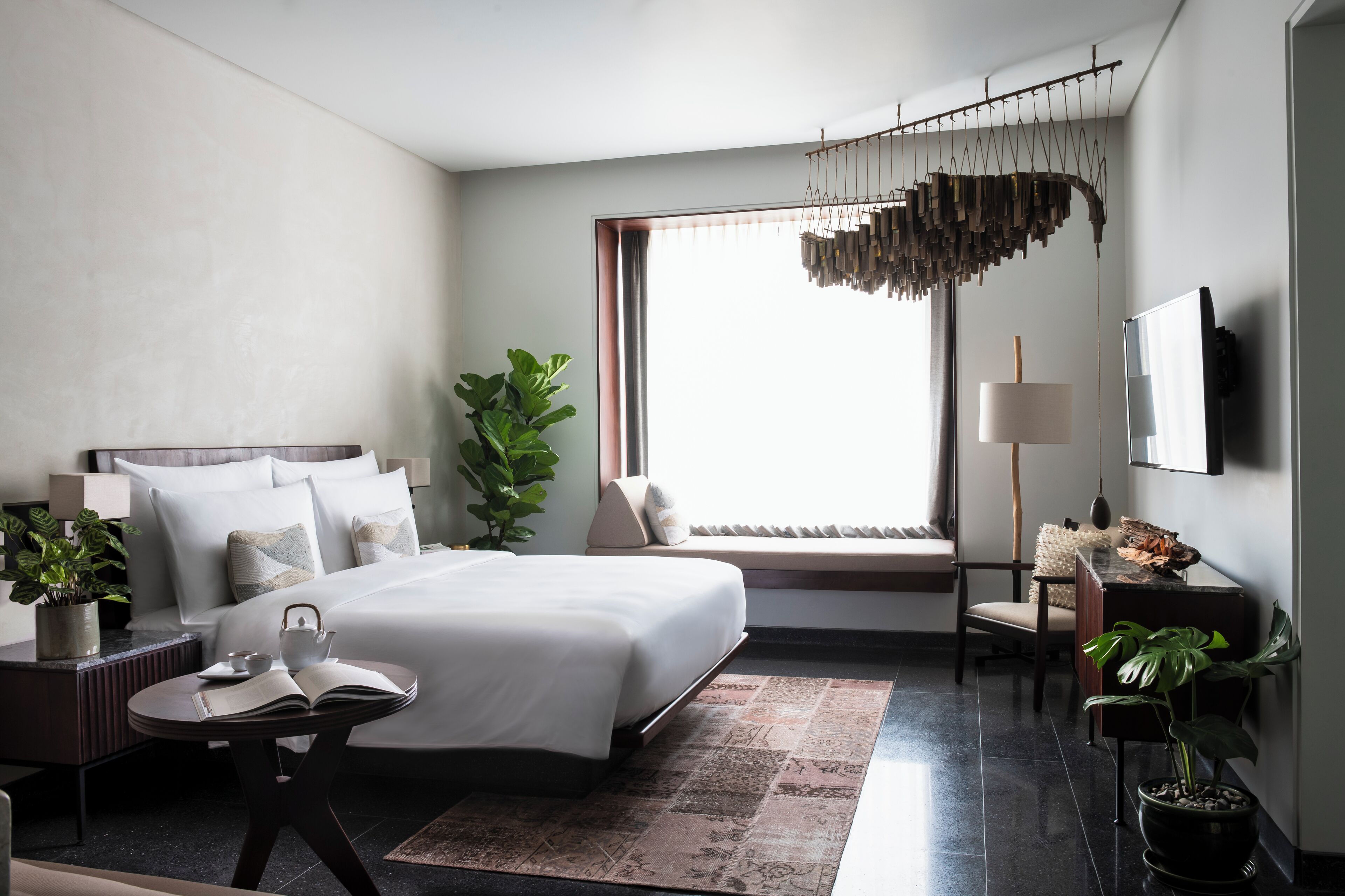 Quarto de hotel moderno com uma cama grande, mesa de cabeceira com um livro e um bule, uma planta em vaso, escrivaninha com luminária e vista para o exterior por uma grande janela.