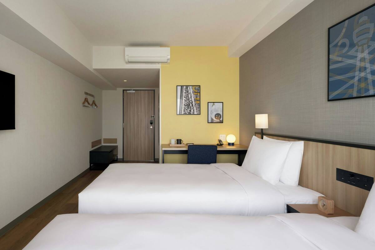 Quarto de hotel com duas camas, uma escrivaninha, uma cadeira, um abajur e uma obra de arte na parede.