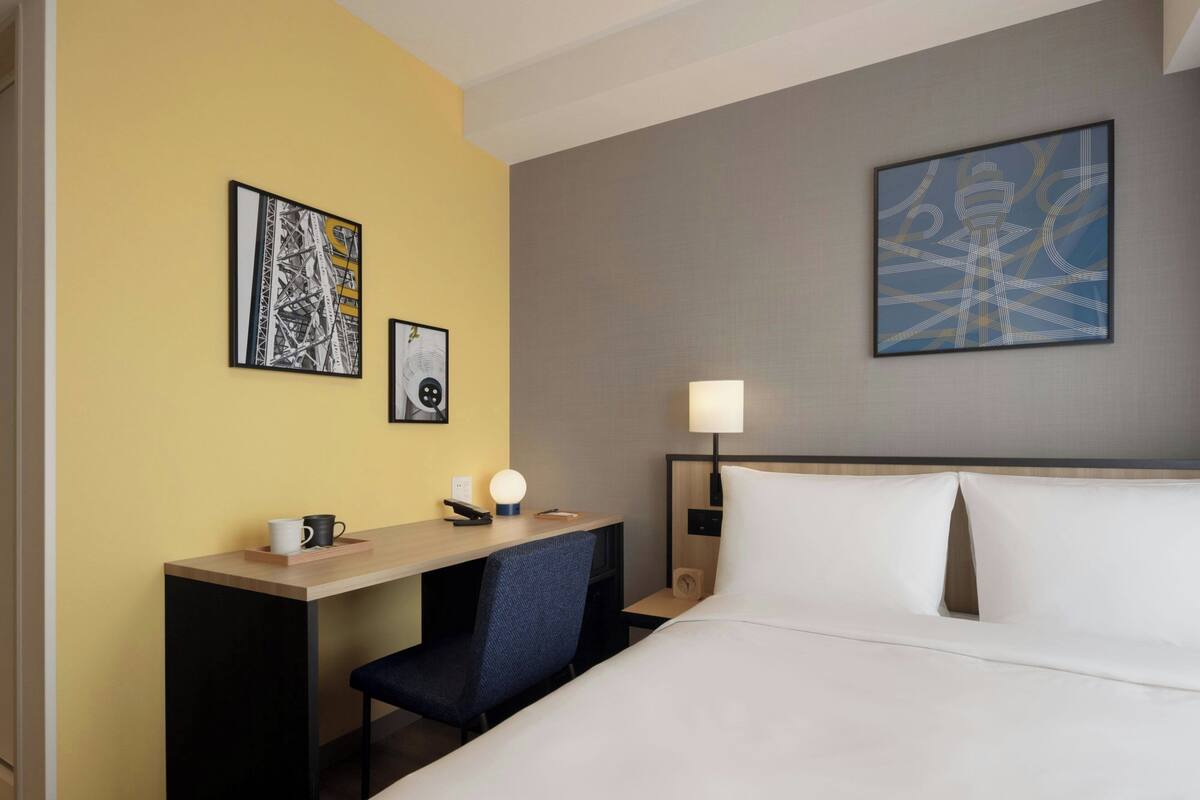 Quarto de hotel com cama, escrivaninha, cadeira, duas imagens emolduradas na parede e abajur.
