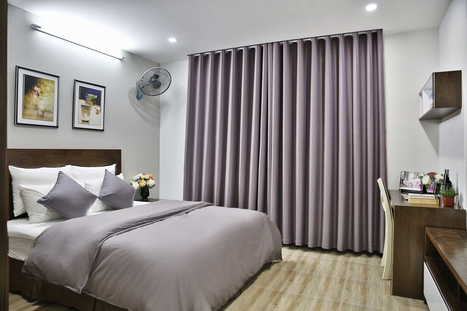 Quarto de hotel moderno com uma cama grande, mesa de cabeceira com abajur, ventilador fixado na parede e uma janela ampla com cortinas.