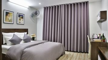 Quarto de hotel moderno com uma cama grande, mesa de cabeceira com abajur, ventilador fixado na parede e uma janela ampla com cortinas.