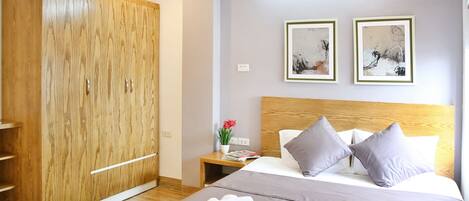 Quarto com guarda-roupa de madeira, cama com roupa de cama cinza, duas pinturas abstratas emolduradas e mesa de cabeceira com um vaso de flores vermelho.