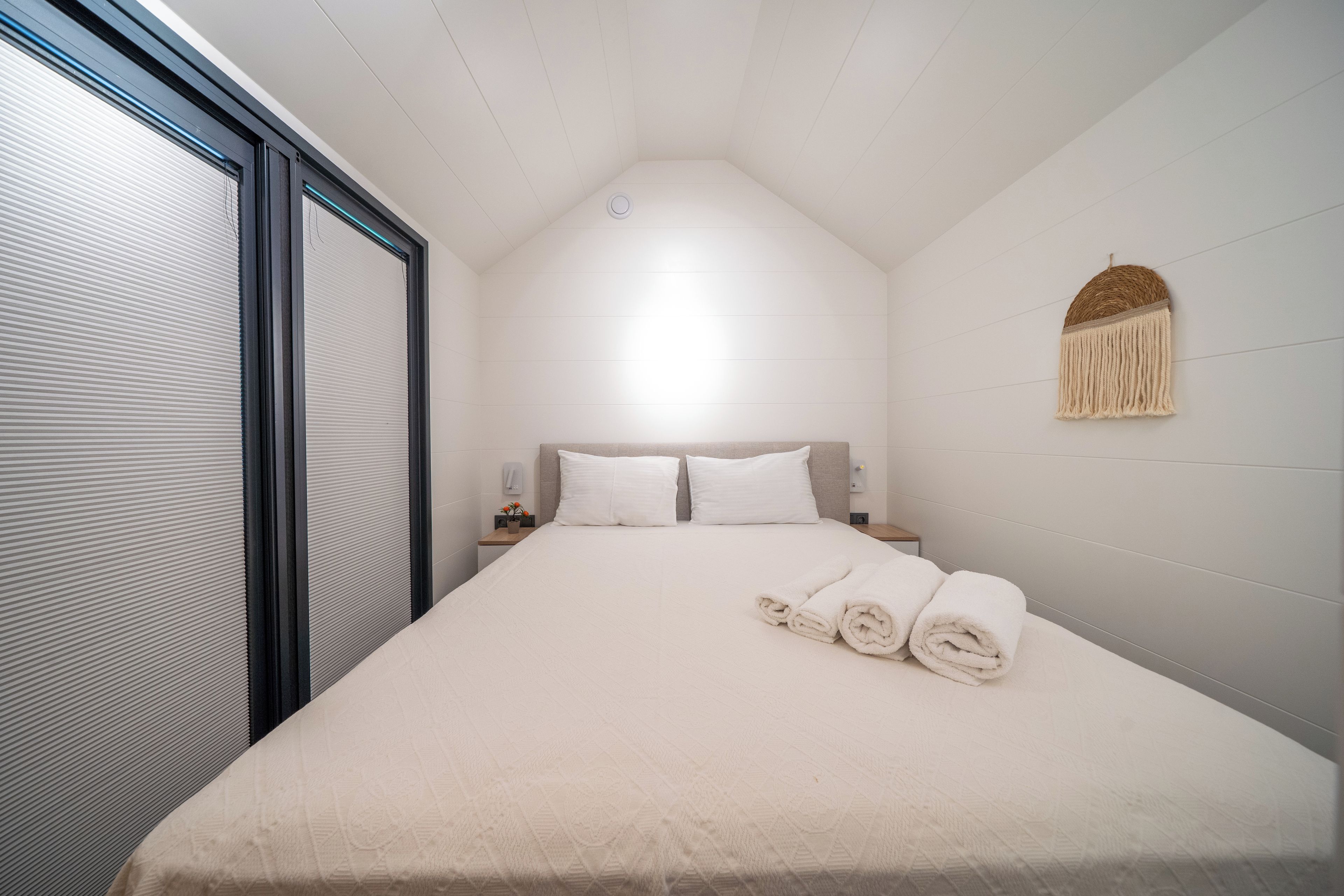 Quarto de hotel com cama, mesa de cabeceira, luminária na parede e janela com persianas.