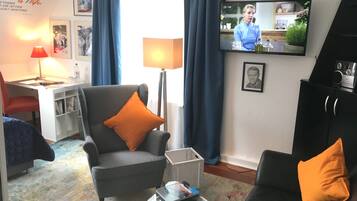 Uma sala de estar com uma poltrona cinza, um sofá preto, uma mesa de centro de vidro e uma televisão fixada na parede.