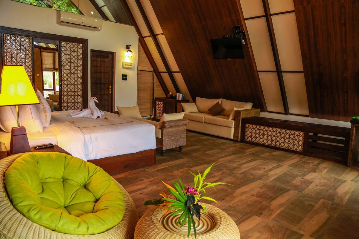 Quarto com cama, sofá, uma poltrona verde, televisão fixada na parede e teto de madeira.