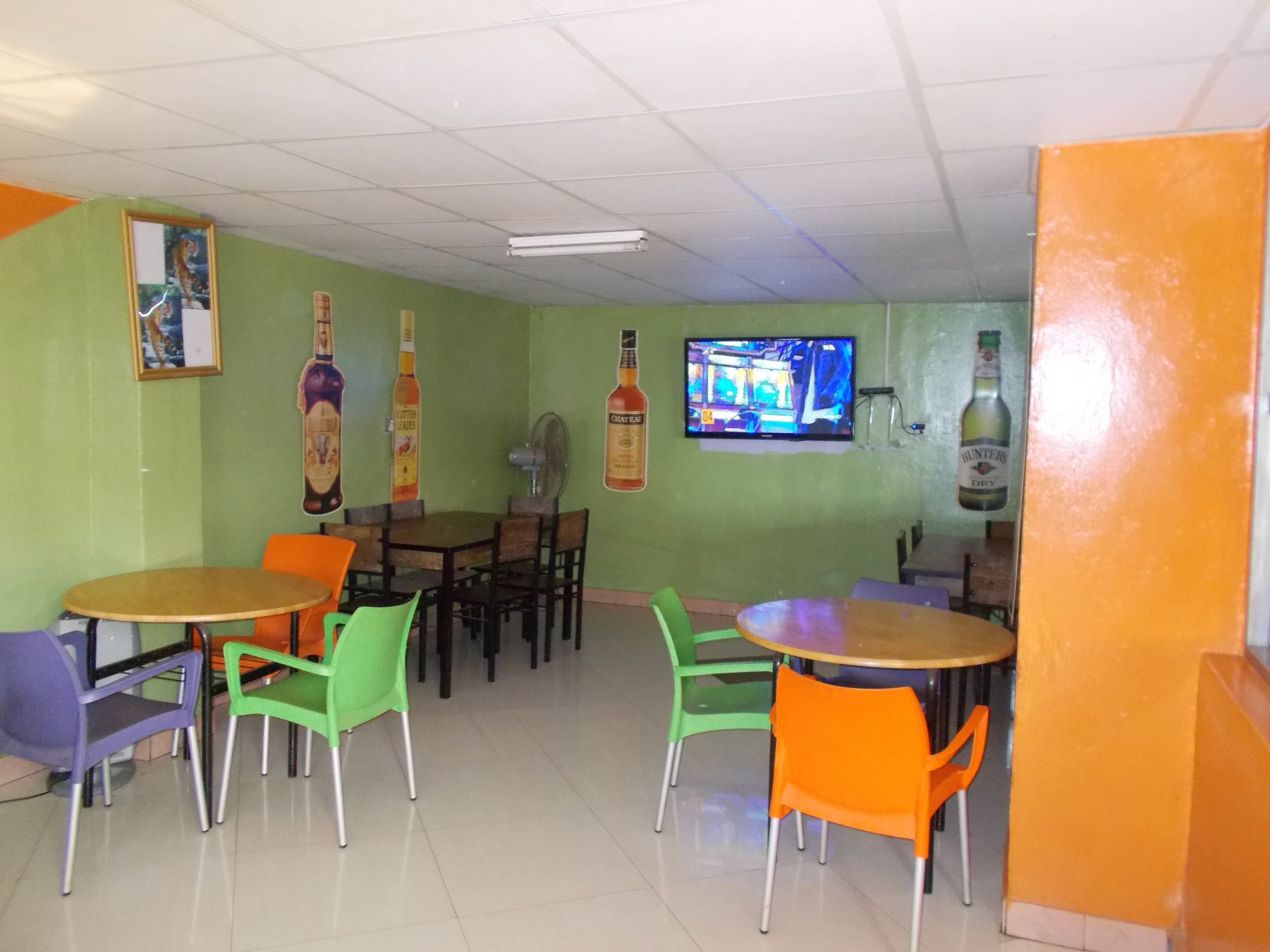 Interior de restaurante com paredes verdes, várias mesas e cadeiras, uma tela de televisão e diversos cartazes de bebidas alcoólicas.
