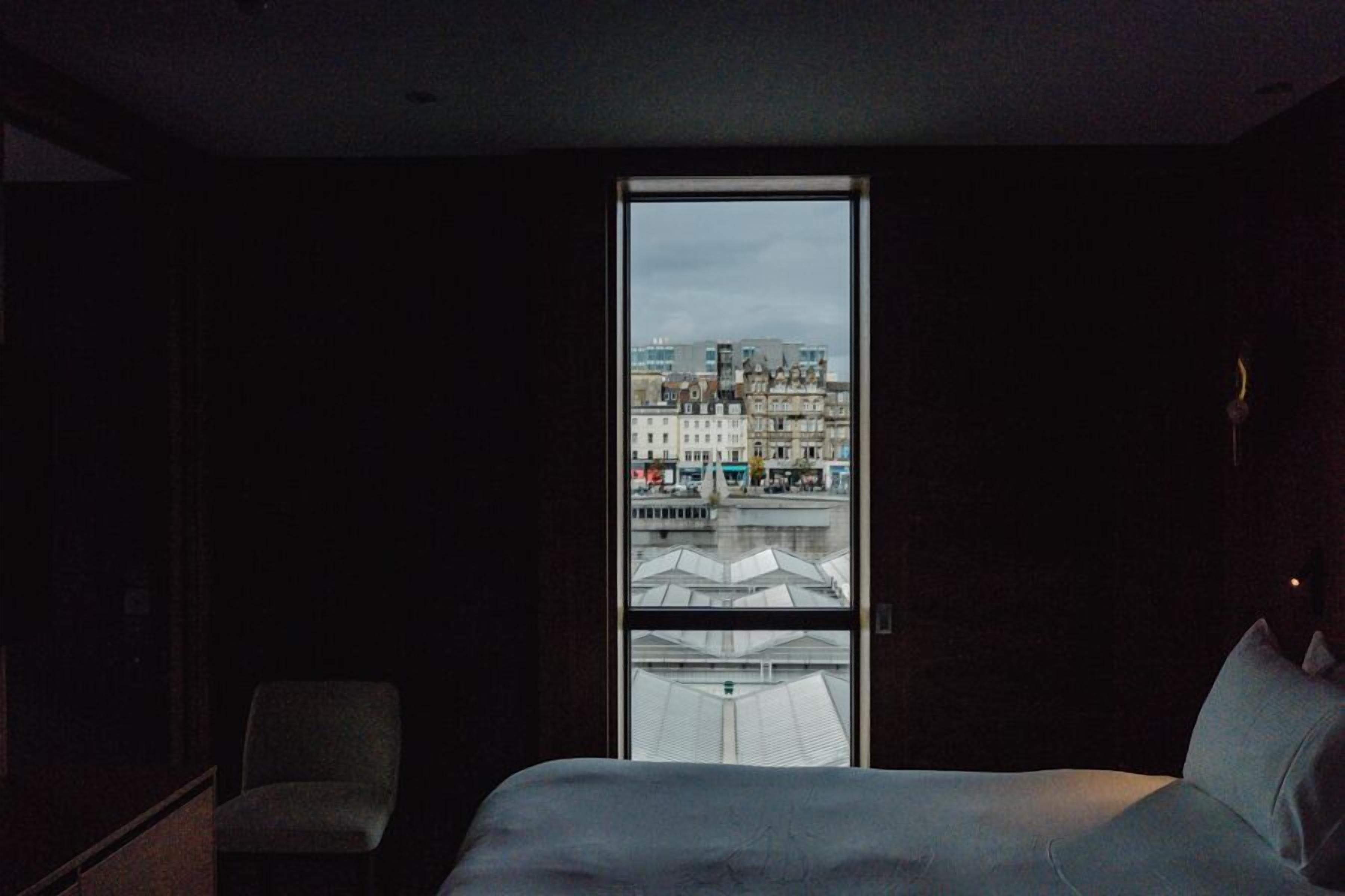 Quarto de hotel com uma cama grande, uma cadeira e vista para a cidade através de uma janela.