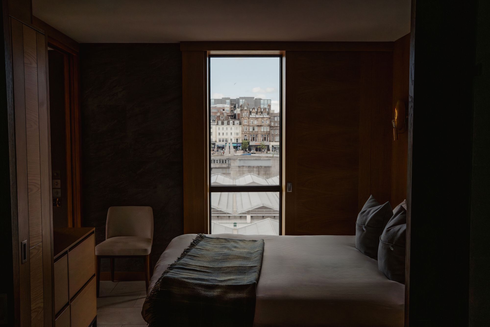 Quarto com cama, cadeira, guarda-roupa e uma janela grande com vista para a cidade.